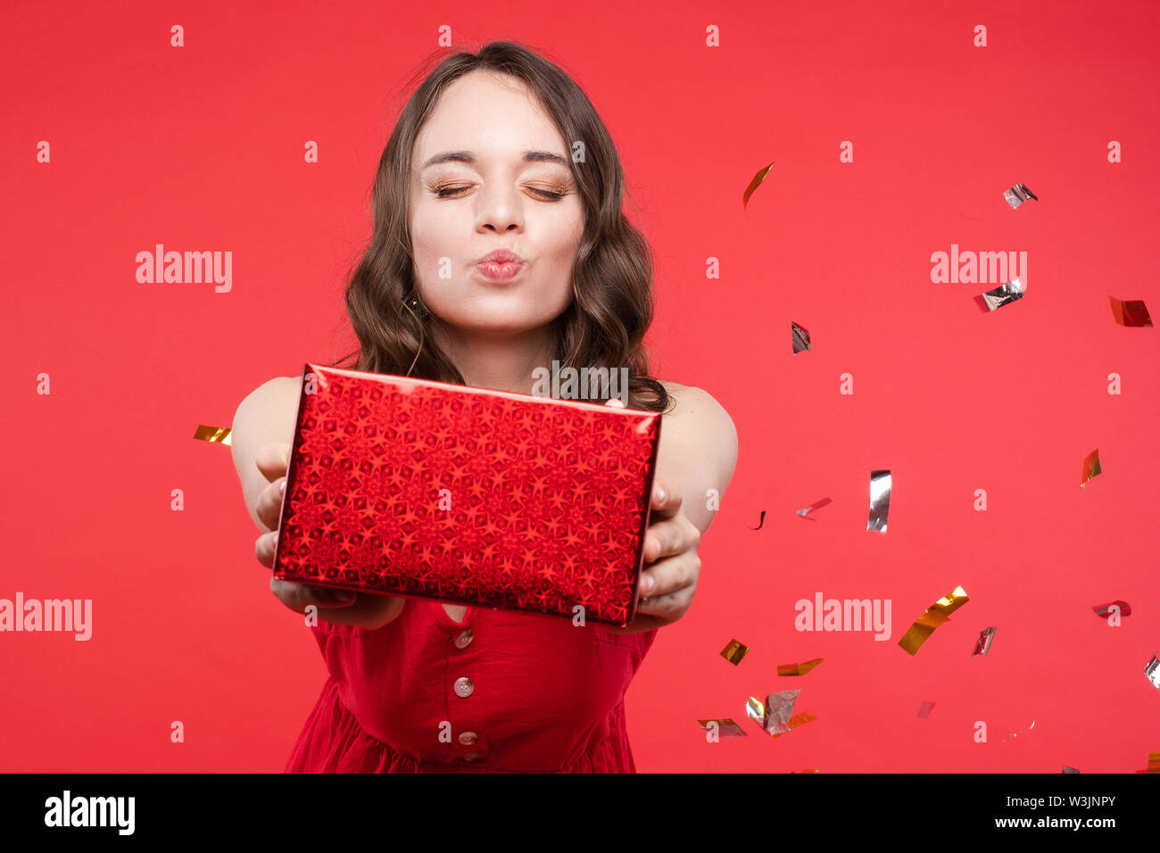 Pleasant young woman taking gift box kissing with closed eyes at red ...