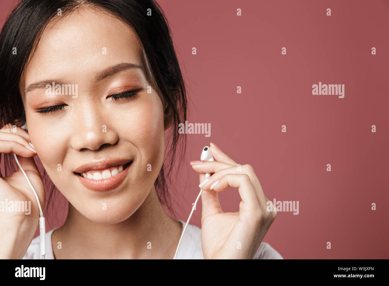 Portrait of joyous asian woman dressed in basic wear smiling and listening to music with ...