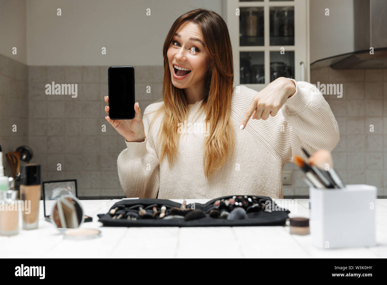 Beautiful young woman doing her makeup at the kitchen, showing blank ...