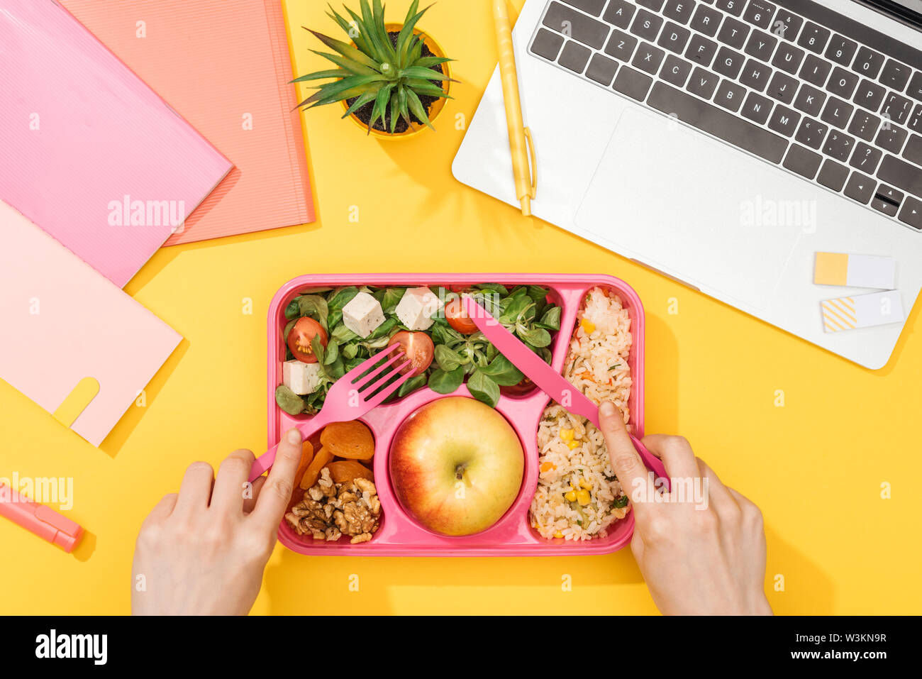 cropped view of woman holding fork over lunch box with food near laptop ...