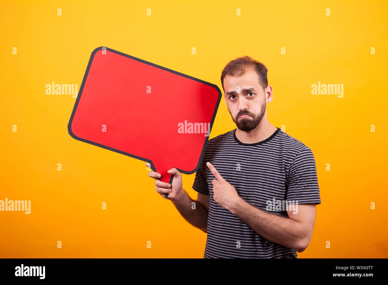 Portrait of sad young man pointing at his informing sign over yellow ...