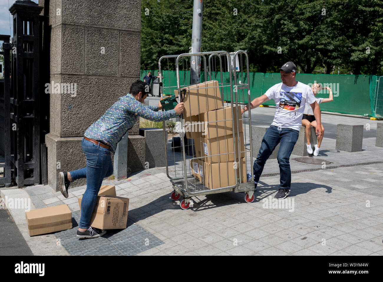 As a young woman poses for photos on the Southbank, deliverymen push a ...