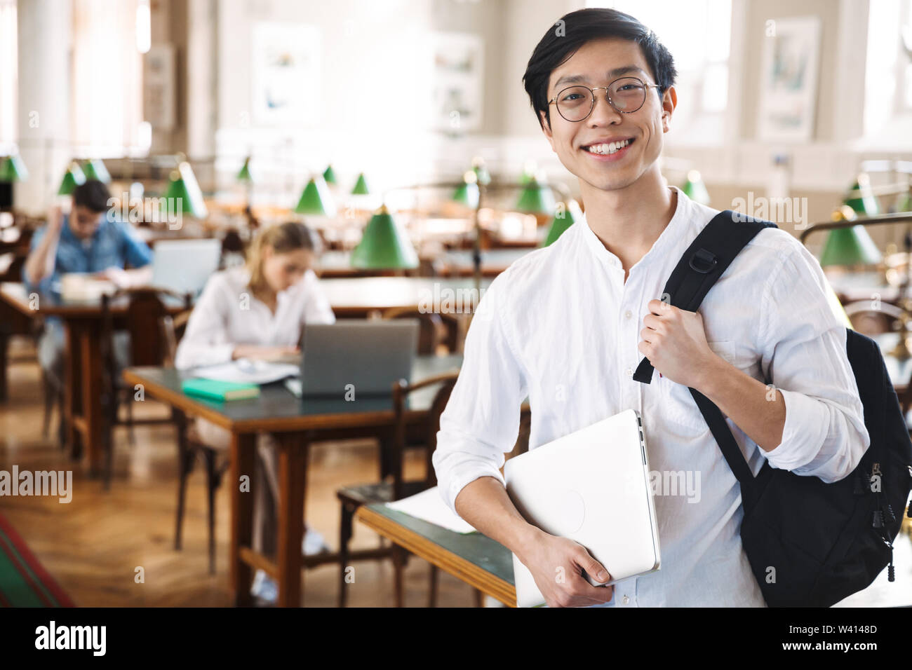 Smiling asian teenager student studying at the library, holding laptop ...