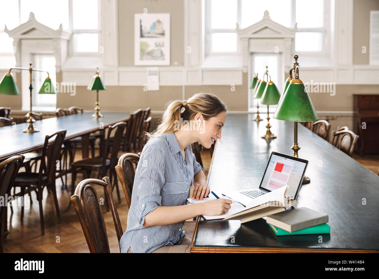 Attractive blonde girl student studying at the library with laptop ...