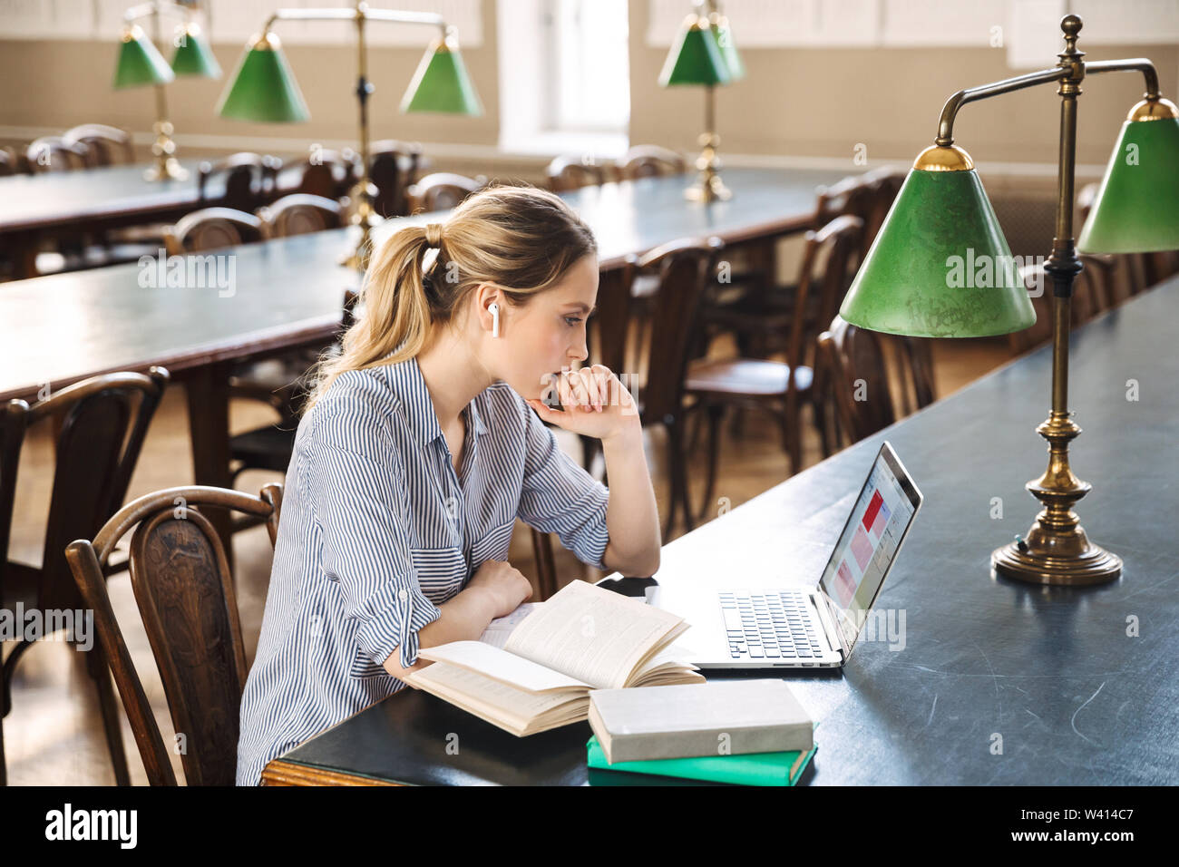 Attractive blonde girl student studying at the library with laptop ...