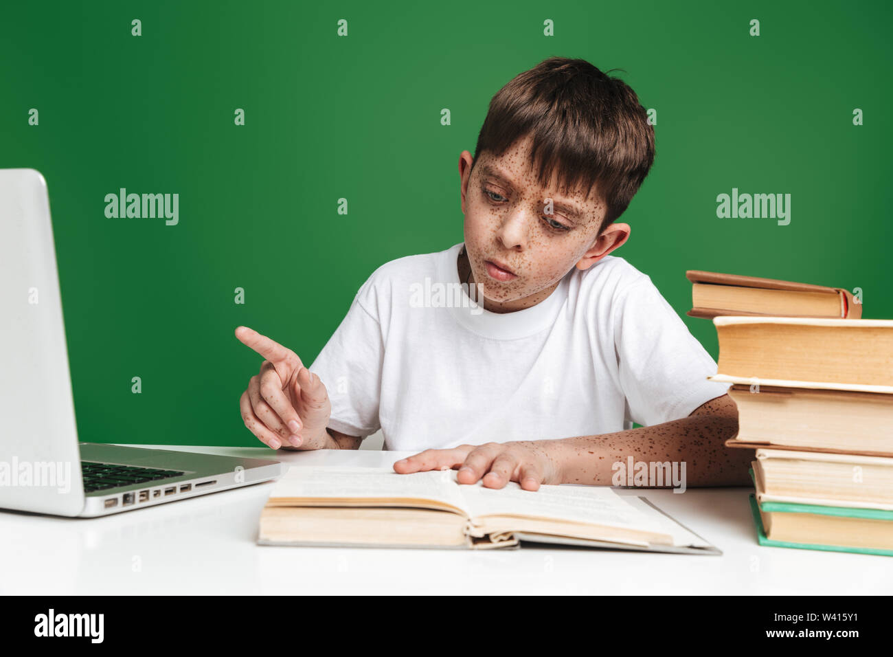 Concentrated young boy with freckles reading book and using laptop ...
