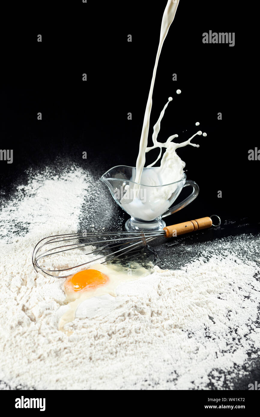 Stream of milk splashing into a pot, a pile of flour and egg on black ...
