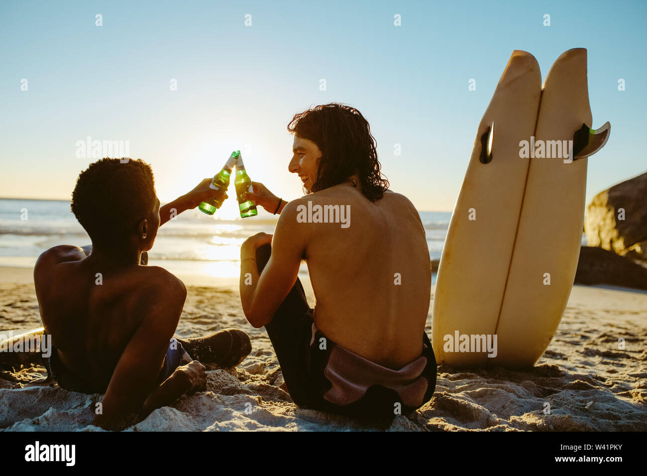 Rear view of two young surfers toasting beers on the beach. Young men ...