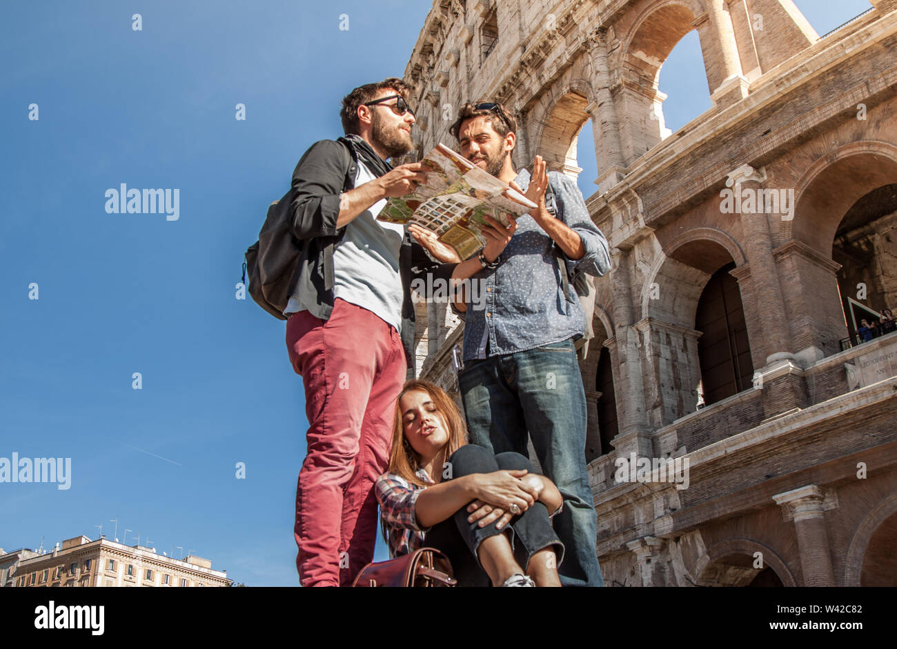 Three happy young friends tourists standing and sitting in front of ...