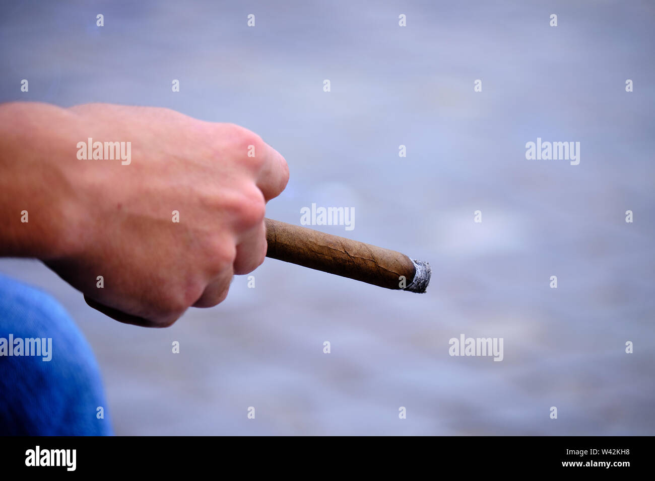 Hand of a young white male holding a lit up cigar while resting hand of ...