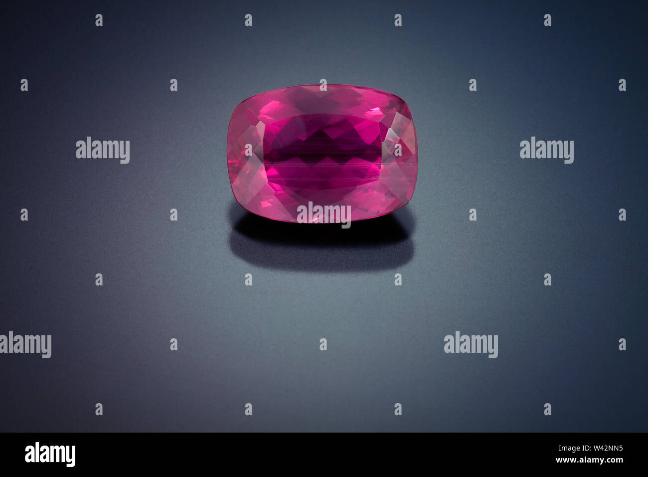 A faceted, rectangle pin cut ruby sits on a reflective black background ...