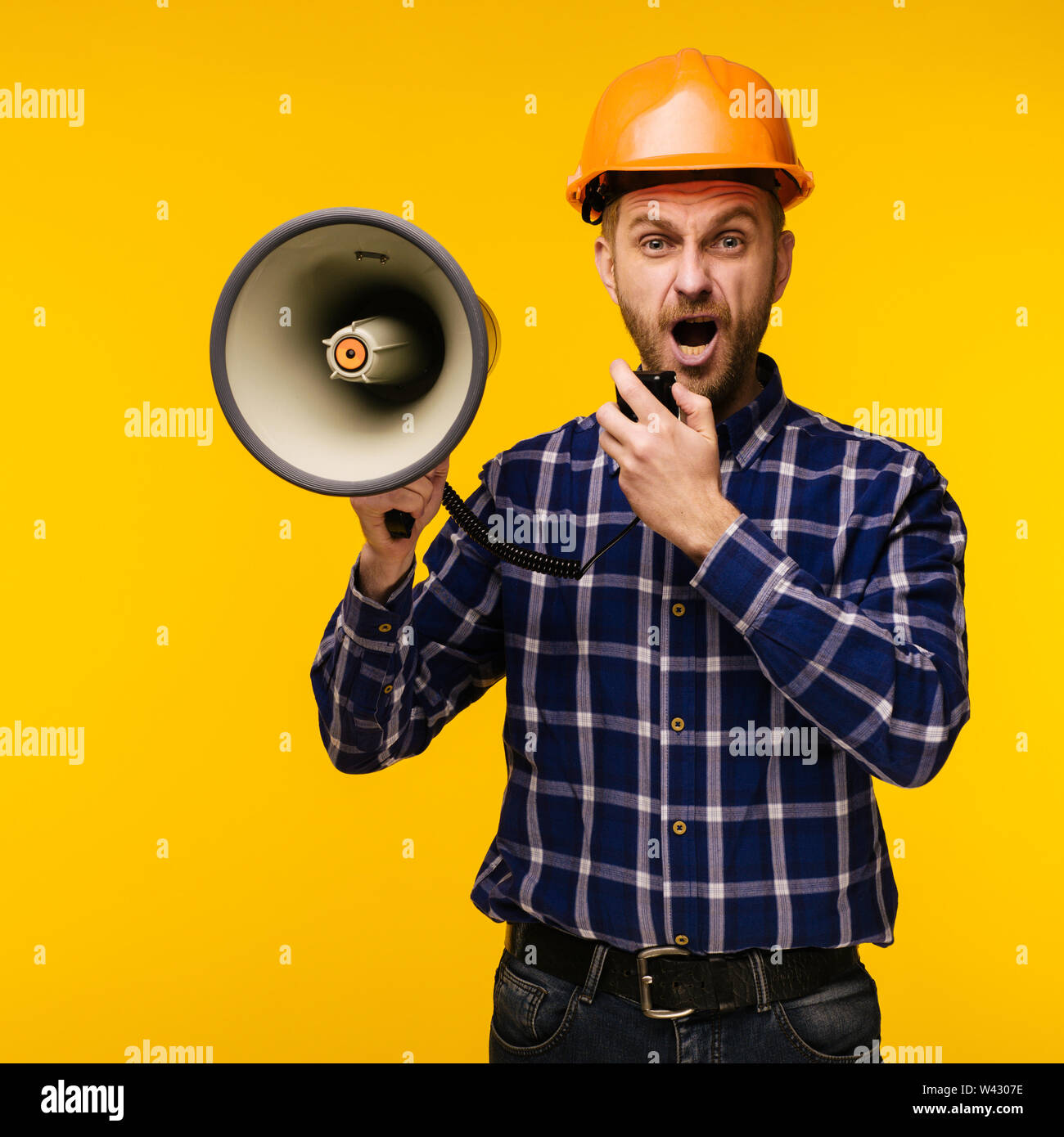 Angry worker man in orange helmet with a megaphone on yellow background ...