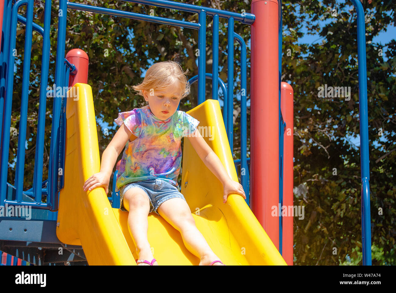 young Caucasian blond girl sliding down a bright yellow playground ...