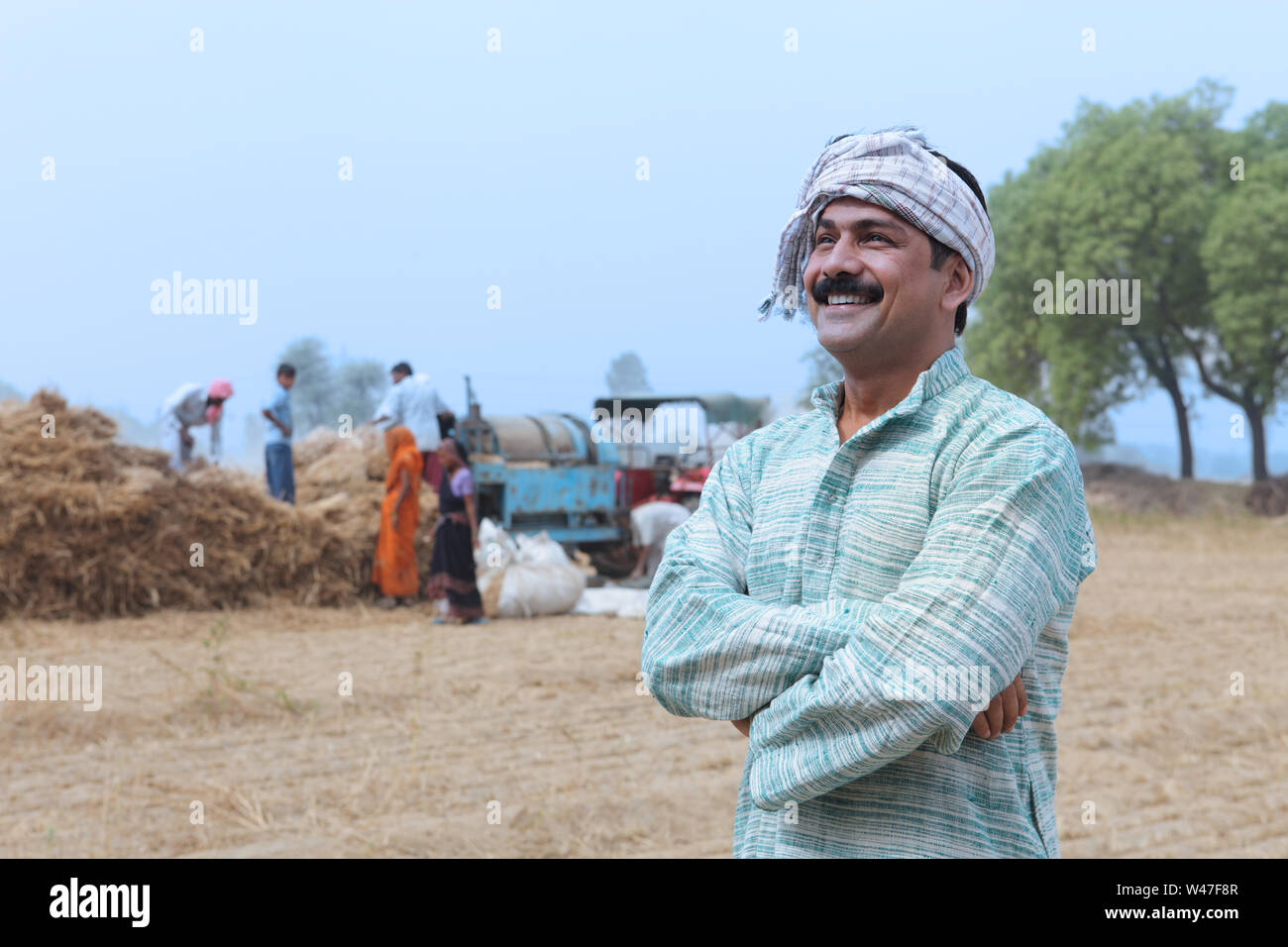 Farmer smiling in a field with farm workers working in the background ...