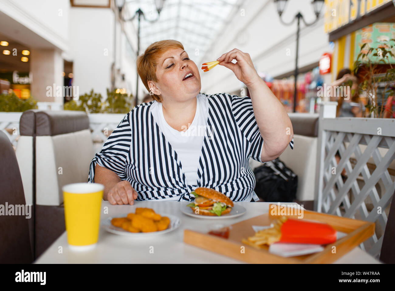 Fat woman eating burger in mall food court. Overweight female person at ...