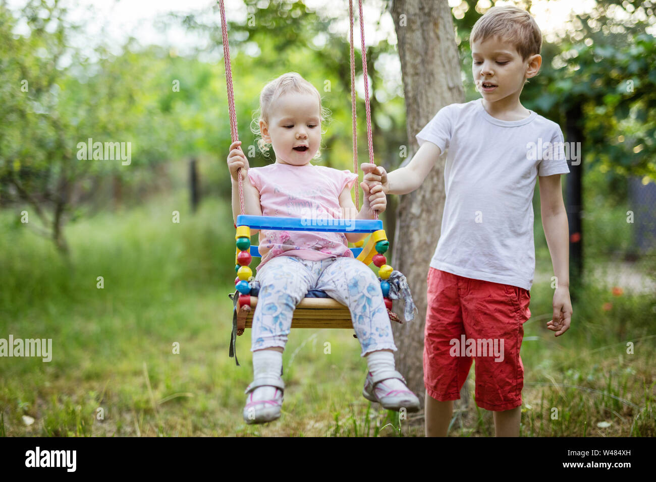Young boy pushing toddler sister on swing. Siblings enjoying summer day ...