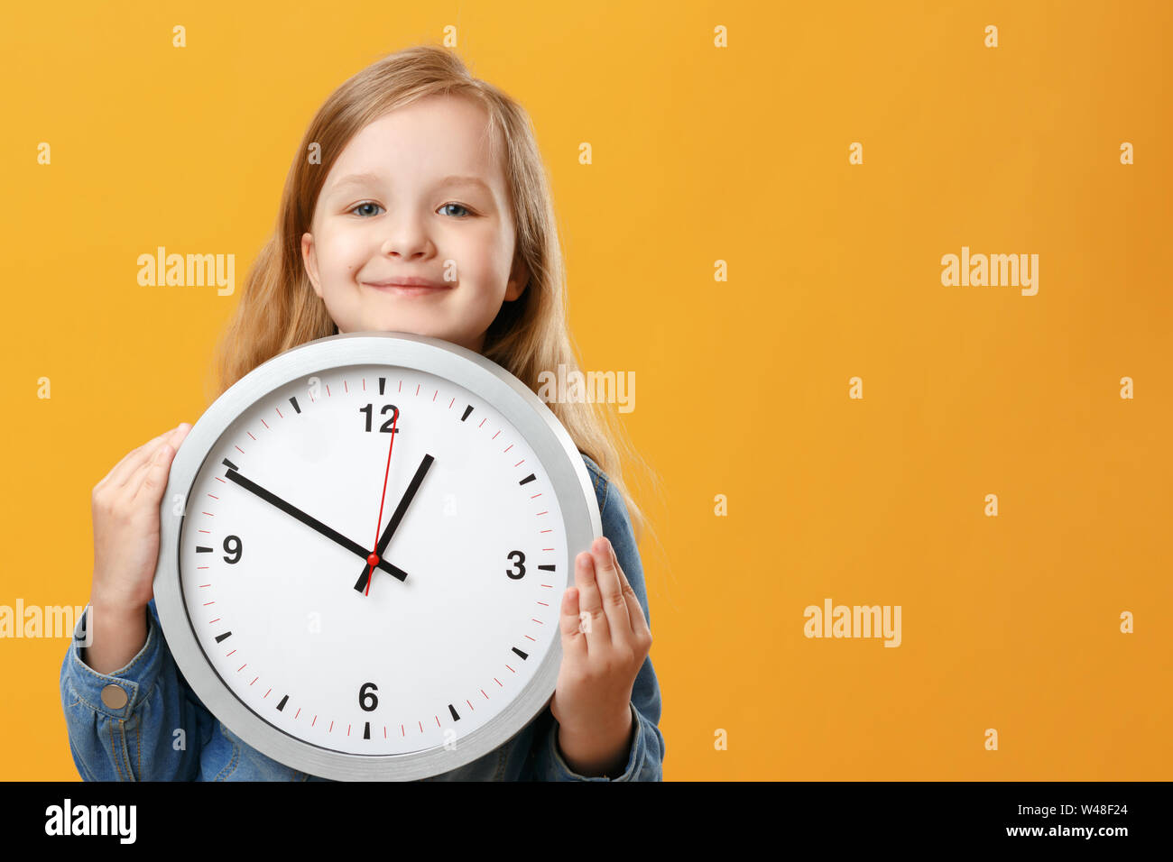 A cute little girl holding a big clock over a yellow background. The ...
