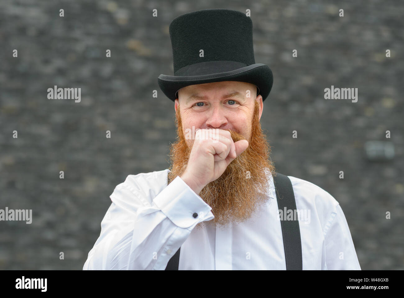 Amused man wearing a top hat and braces giggling behind his hand as he ...