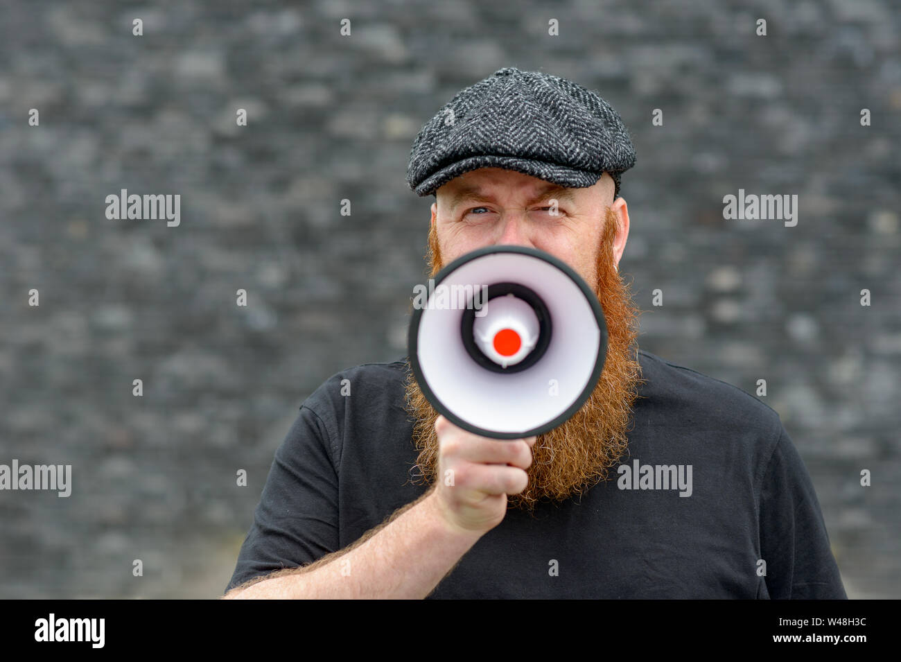 Bearded man wearing a cloth cap speaking into a megaphone making an ...