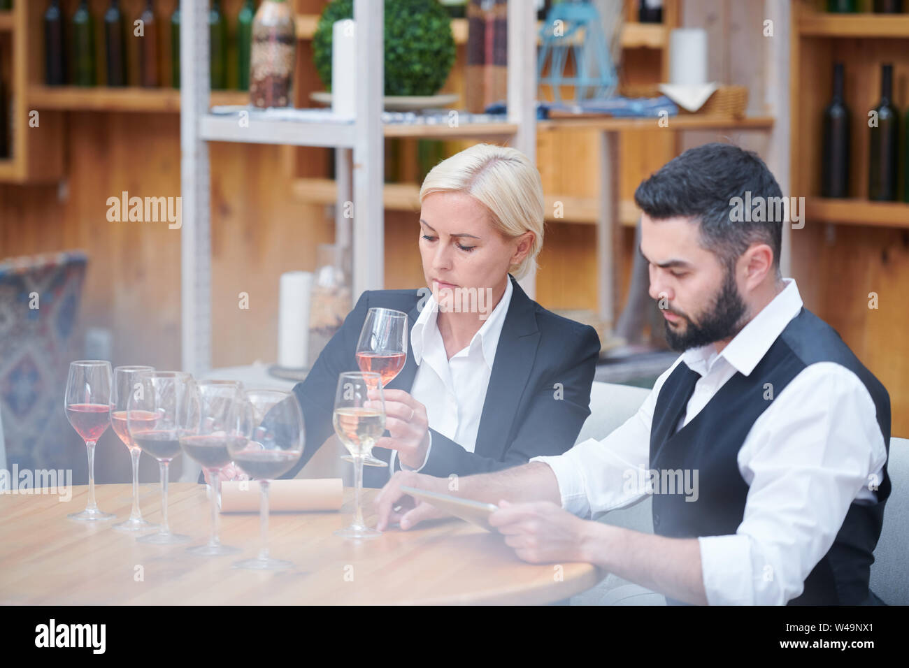 Two co-working sommeliers sitting by table with group of wine samples ...