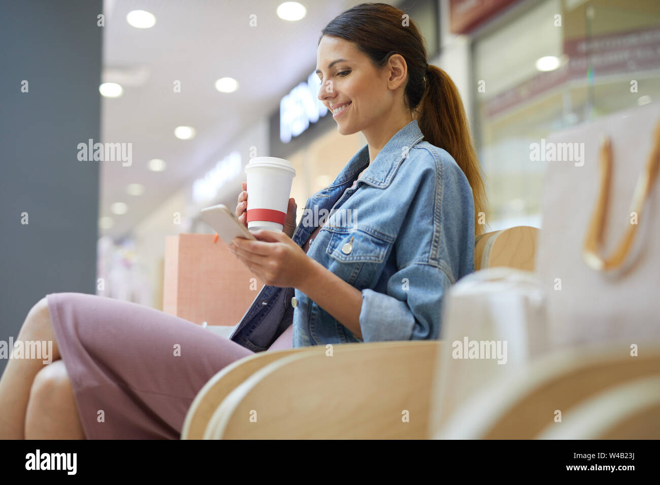 Young attractive girl chilling with coffee in mall and scrolling news feed on her smartphone ...