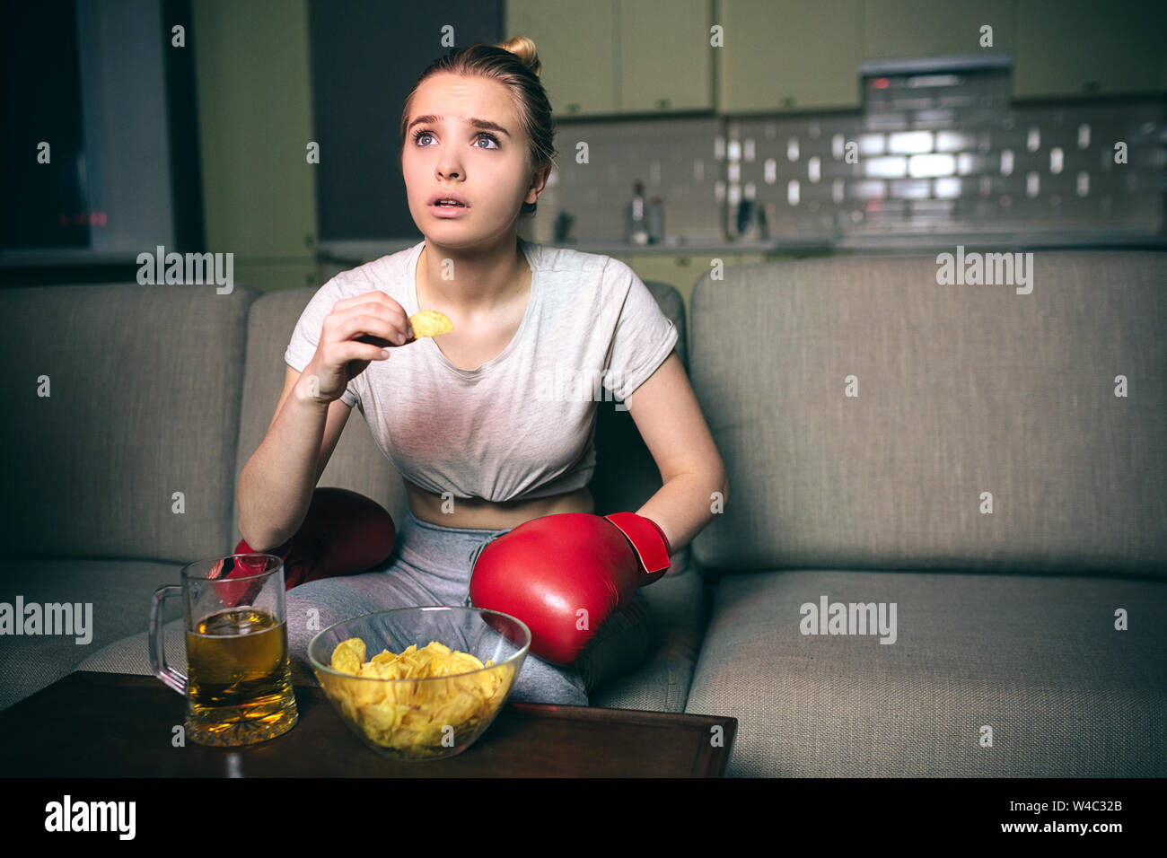 Young woman watch boxing on tv at night. Amazed blonde look up and eat ...