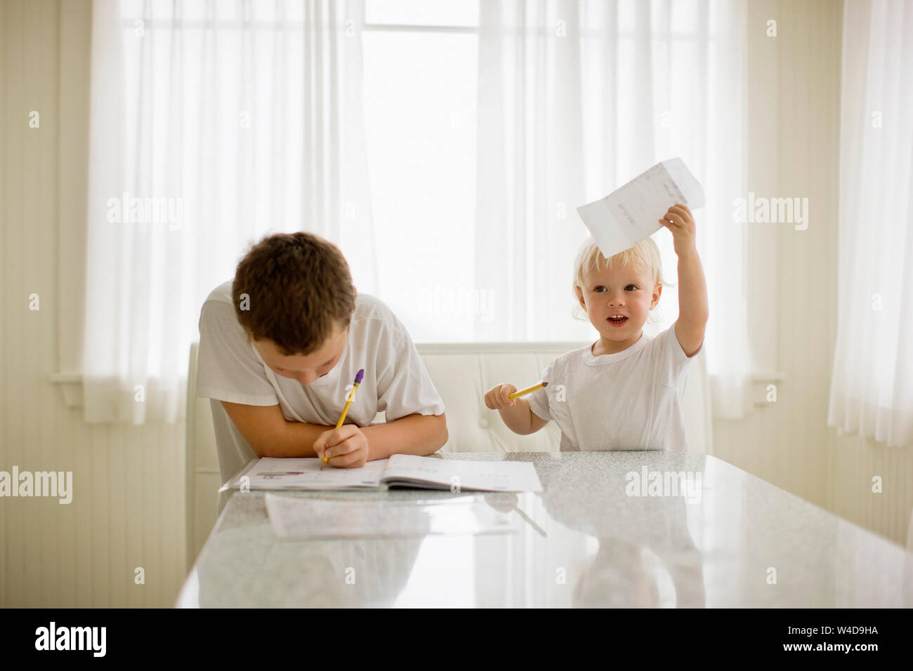 Young boy sitting at a dining table doing his homework with his younger ...