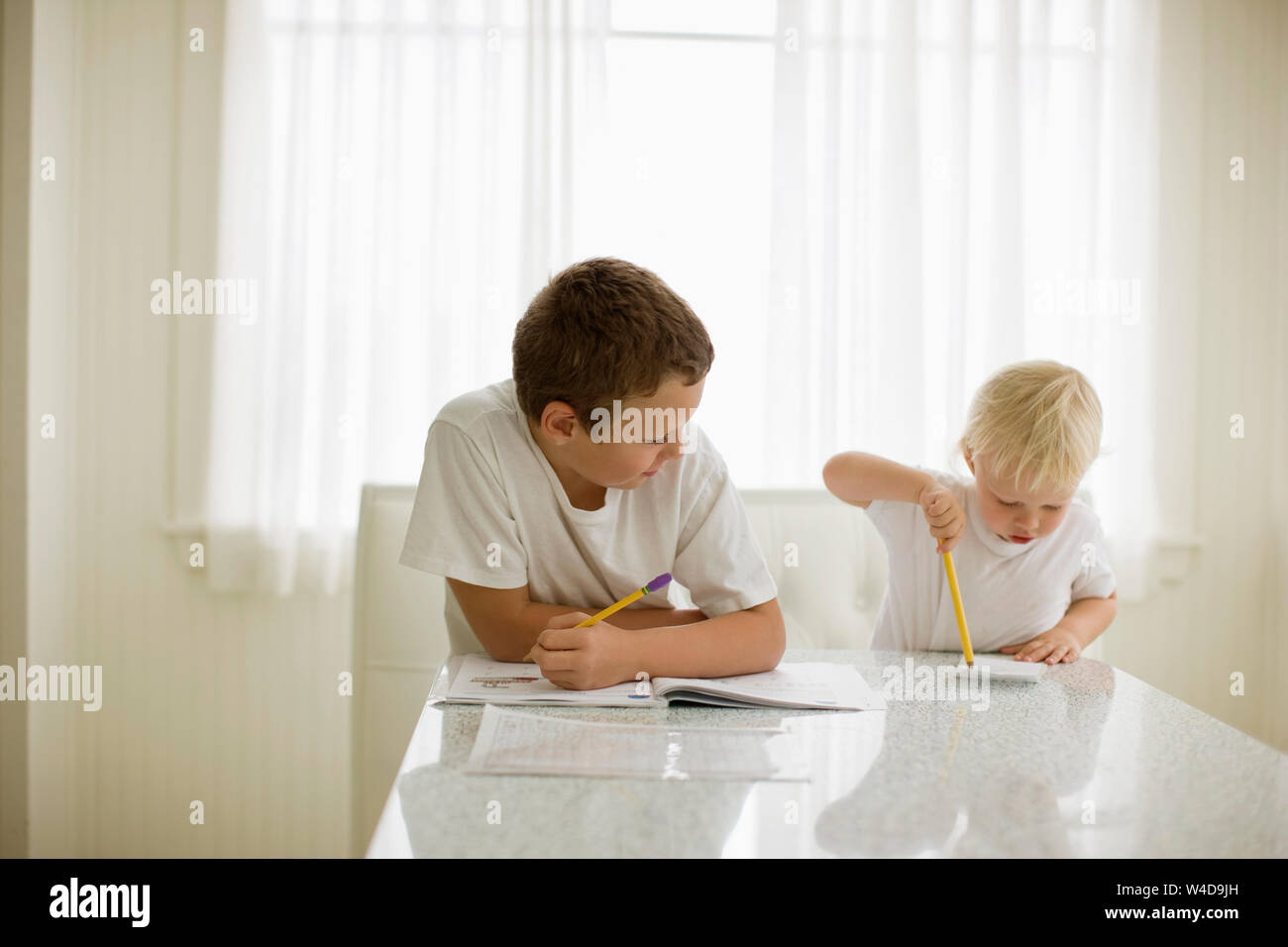 Young boy sitting at a dining table doing his homework with his younger ...