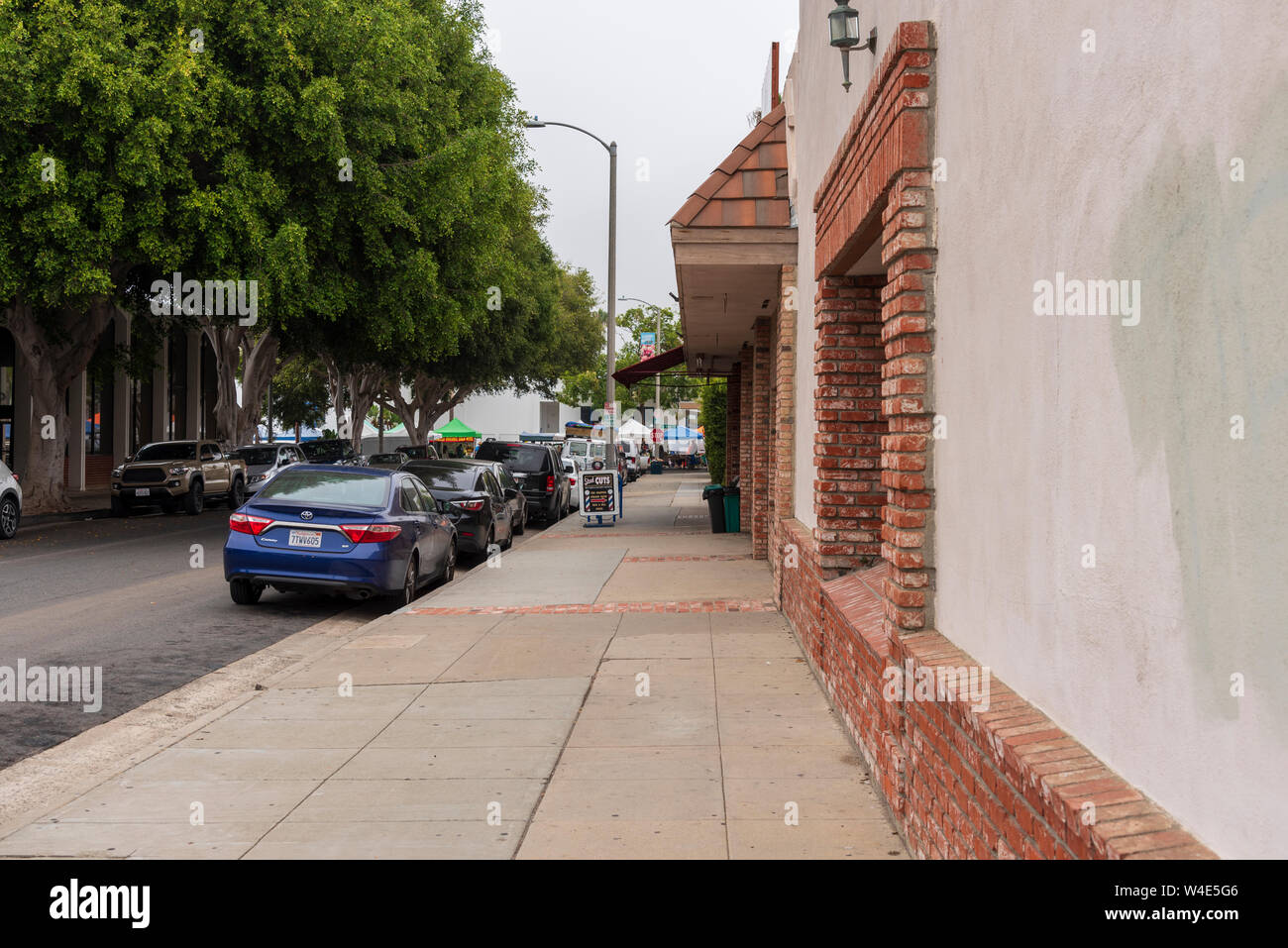 Small town side street with parked cars and buildings with tall green ...