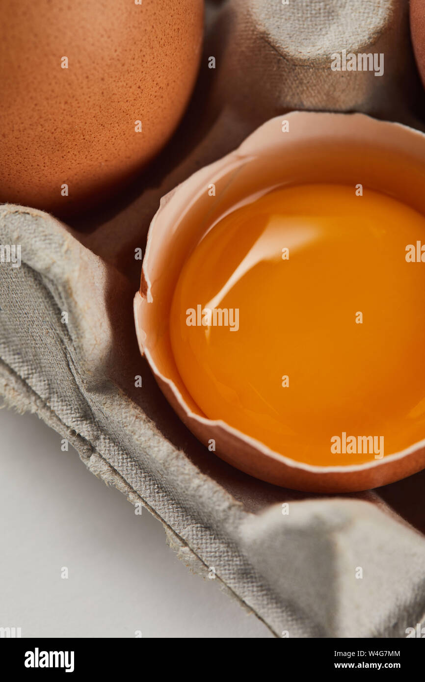 close up view of broken eggshell with yellow yolk near eggs in carton ...