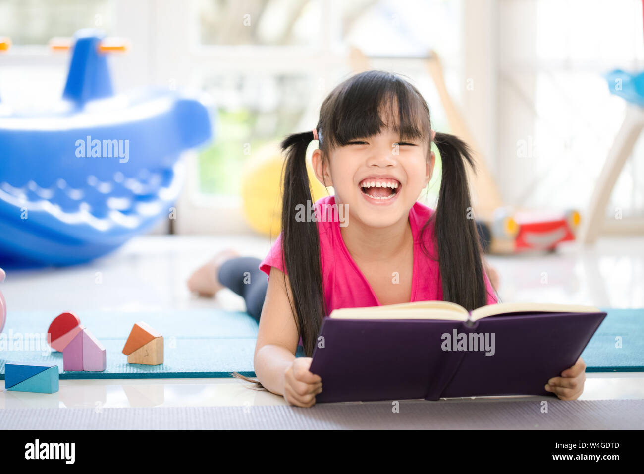 Cute little girl is reading a book . Funny kid having fun in kids room ...