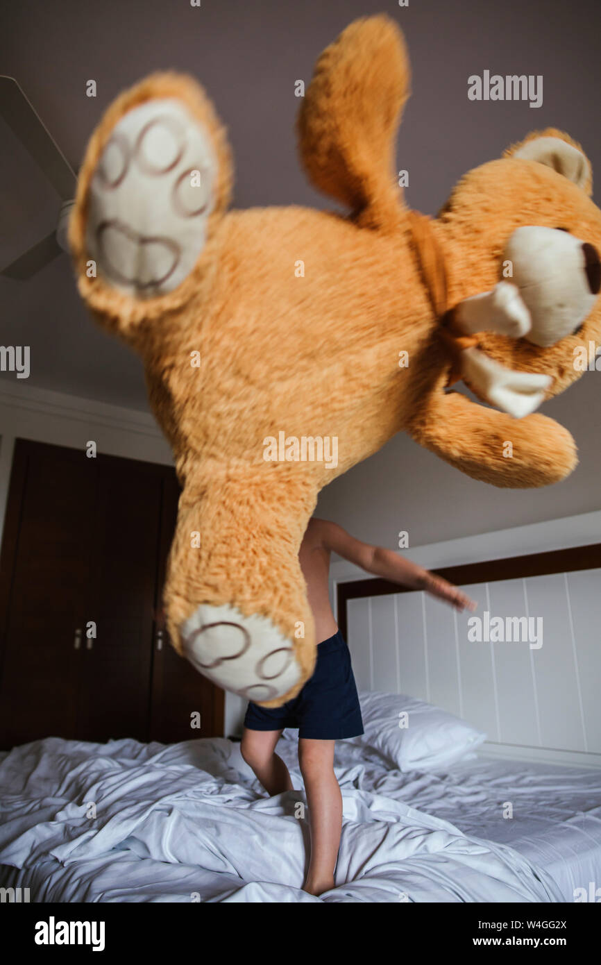 Little boy standing on bed throwing oversized teddy bear in the air ...