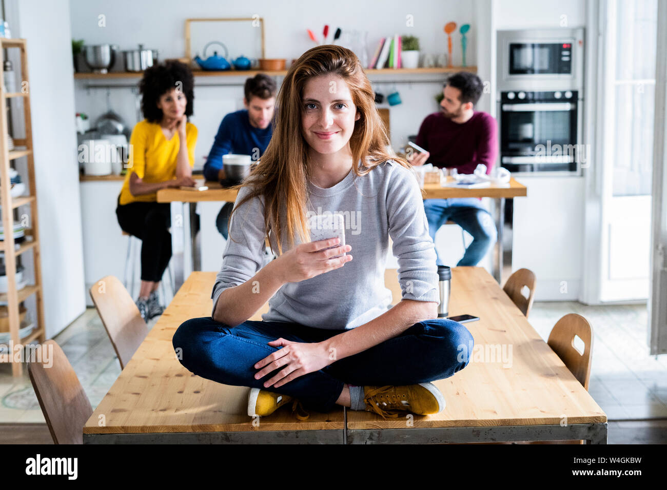 Woman with cell phone sitting on dining table at home with friends in ...
