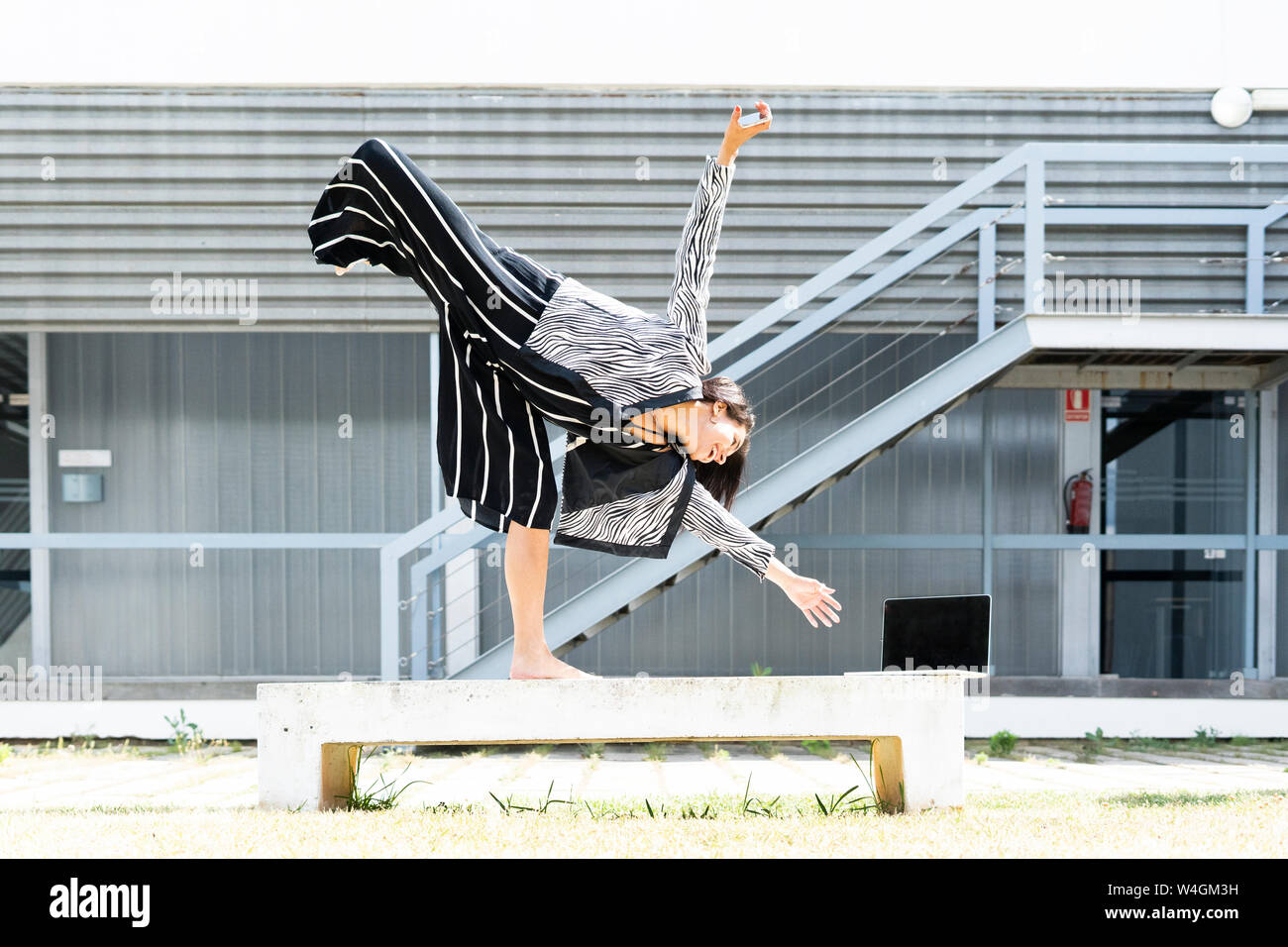 Female ballet dancer lifting her leg on a bench in front of an office ...