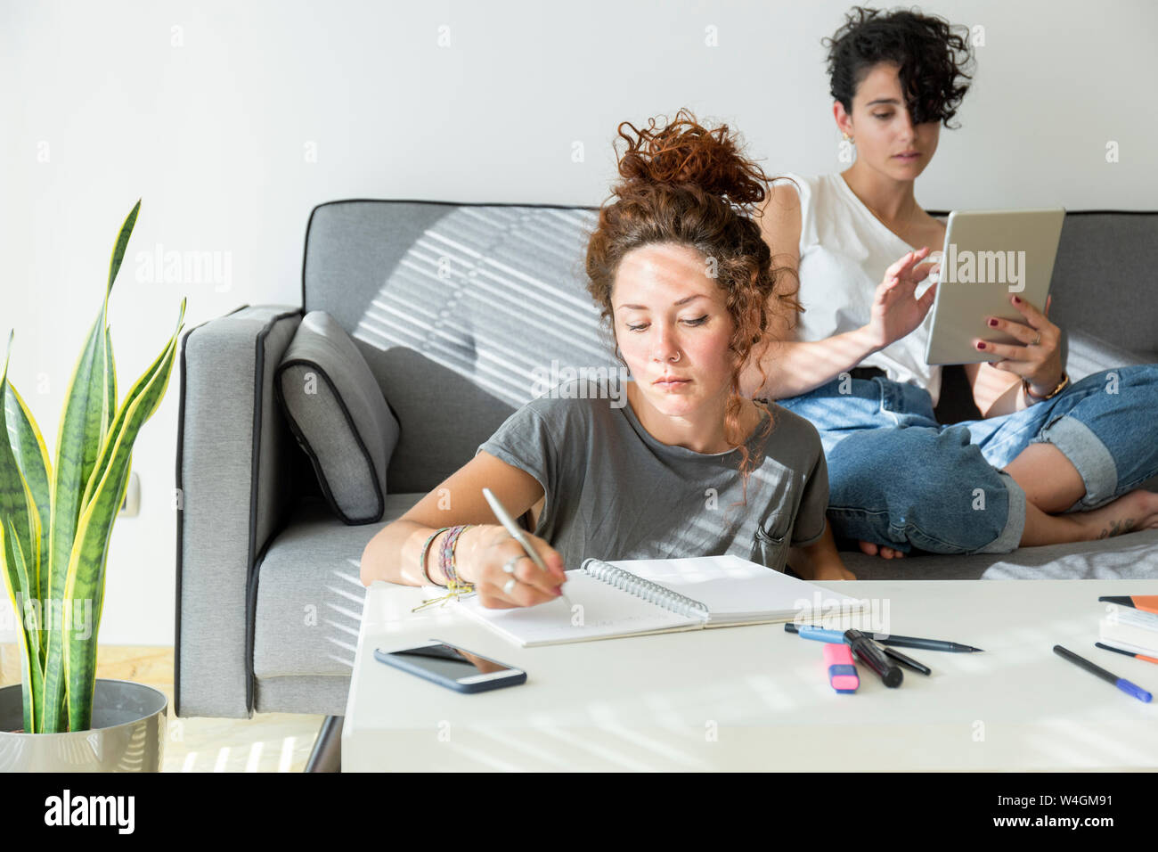 Young woman taking notes at home with friend sitting on couch using ...