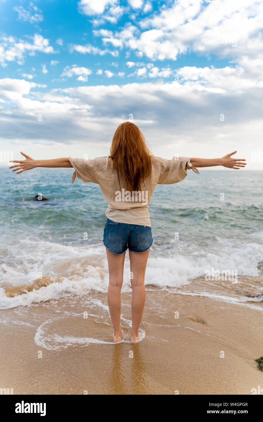 Back view of redheaded young woman standing at seafront with arms ...