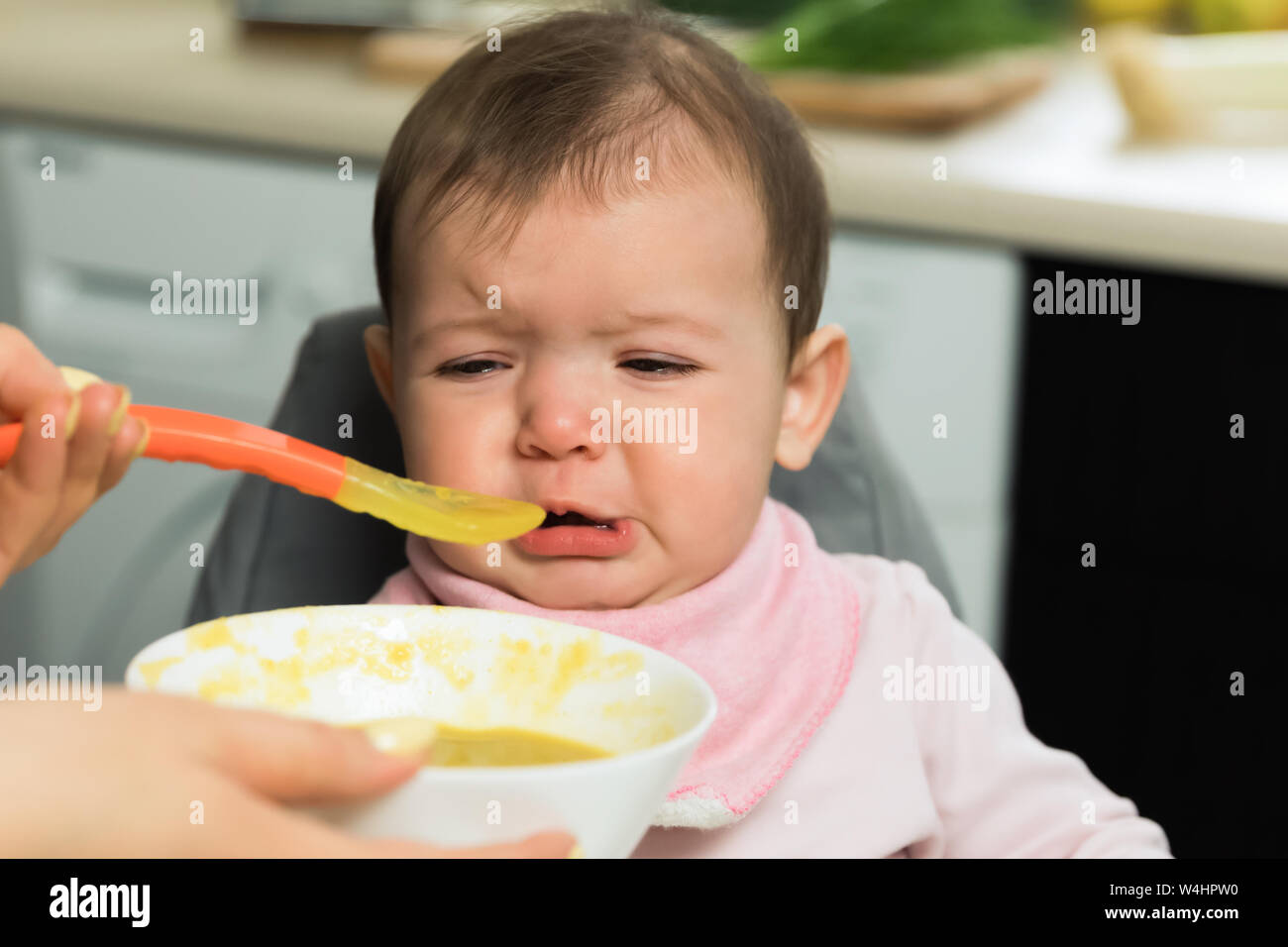 Mom feeds a baby with a spoon. The baby is crying while sitting at the ...