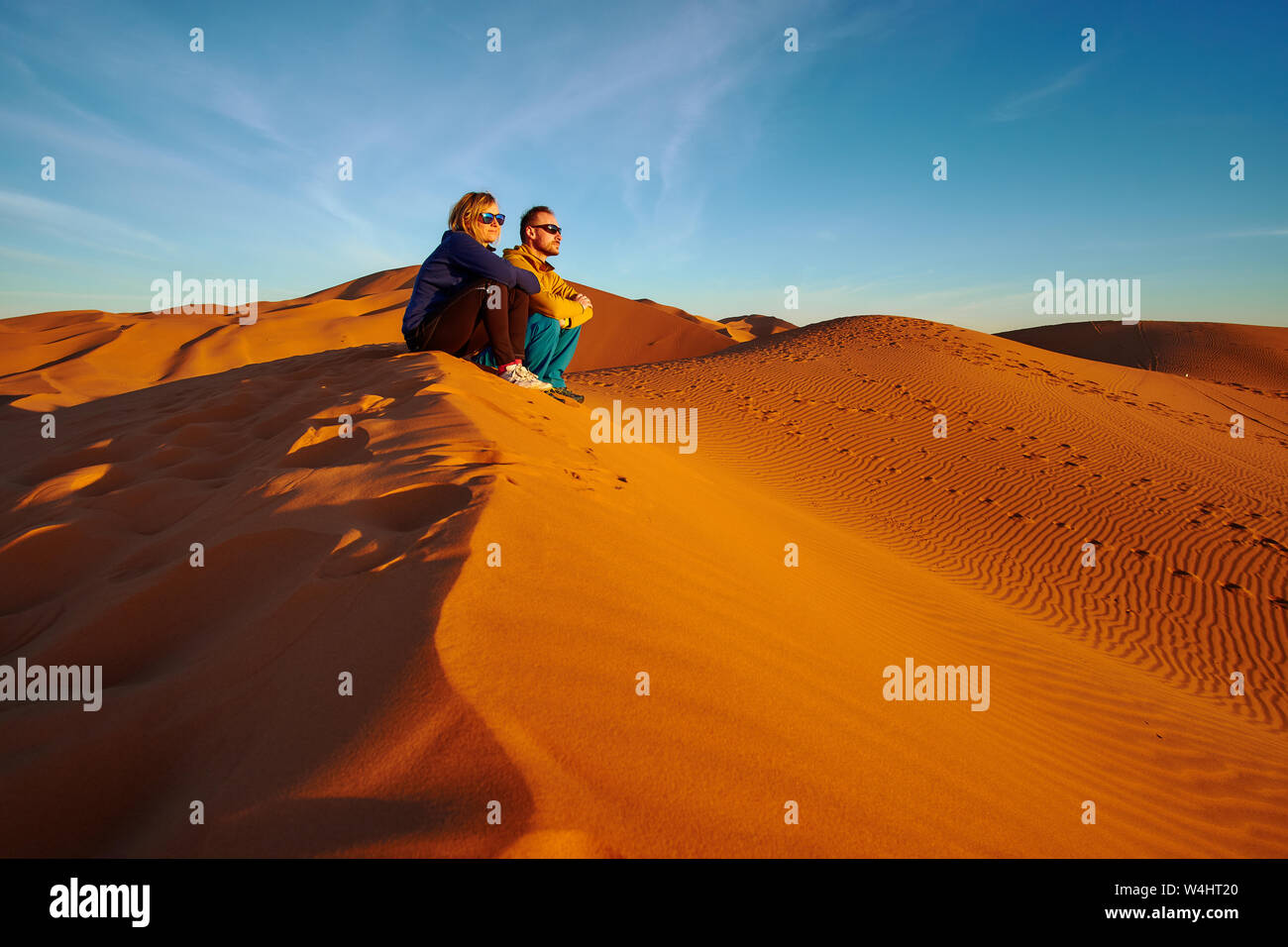 Tourist couple watching sunrise in the desert sitting on a sand dune in ...