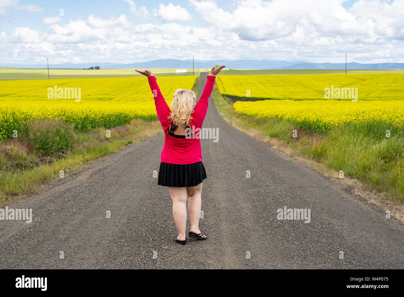 Blonde woman poses on an empty farm road with arms raised, near a field ...