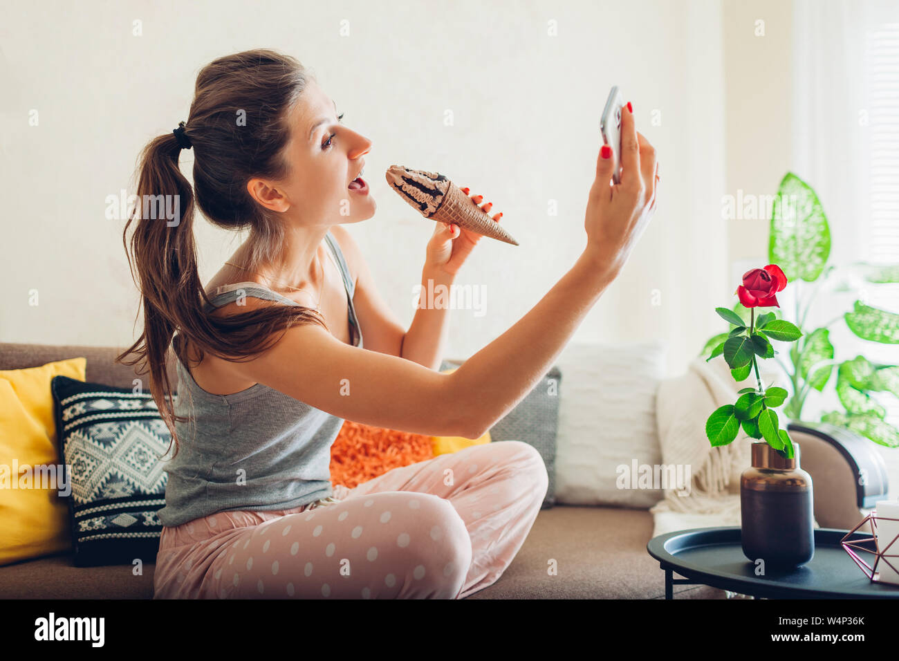 Happy young woman eating chocolate ice-cream in cone sitting on couch ...