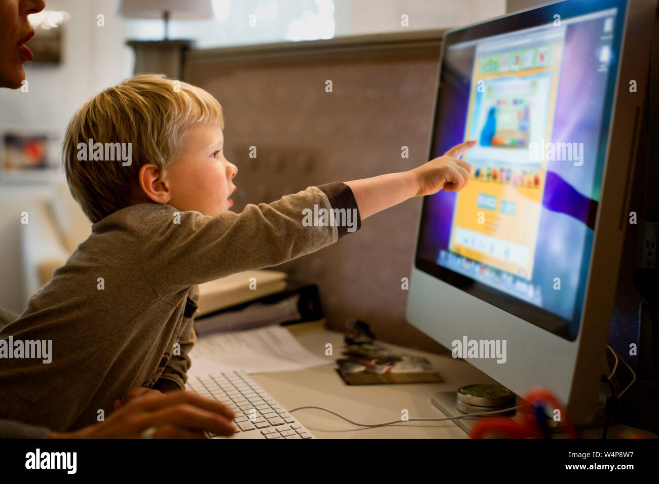 Young boy leans across a desk to point at a large desktop computer ...