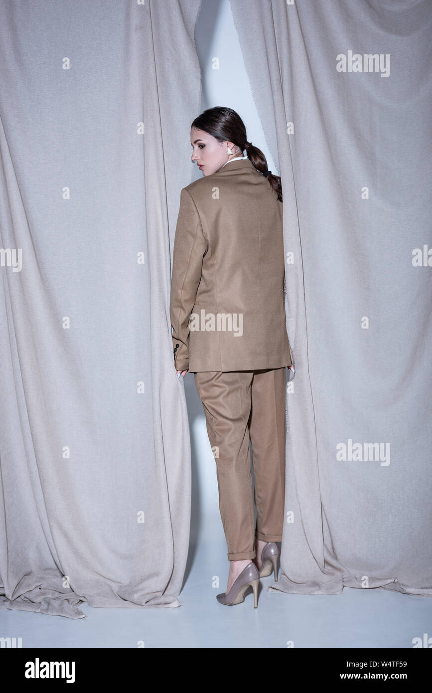 back view of fashionable model in suit standing on light grey curtain ...
