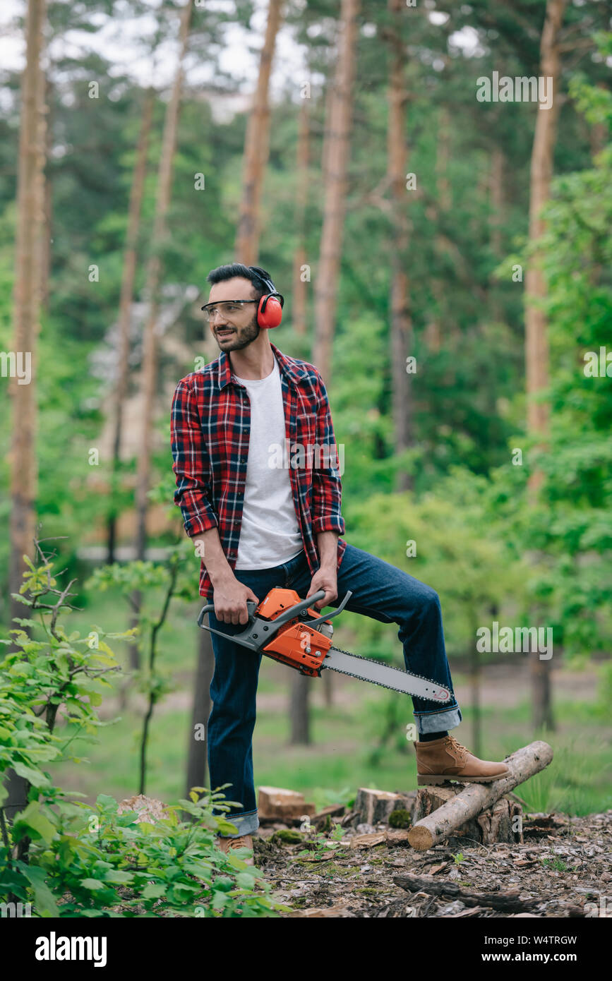 smiling lumberman in plaid shirt and denim jeans standing with chainsaw ...