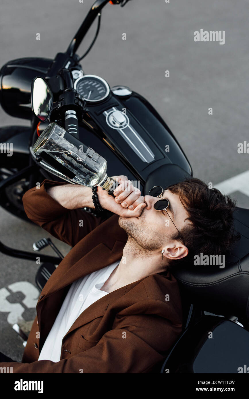 stylish motorcyclist leaning head on motorcycle and drinking alcohol ...