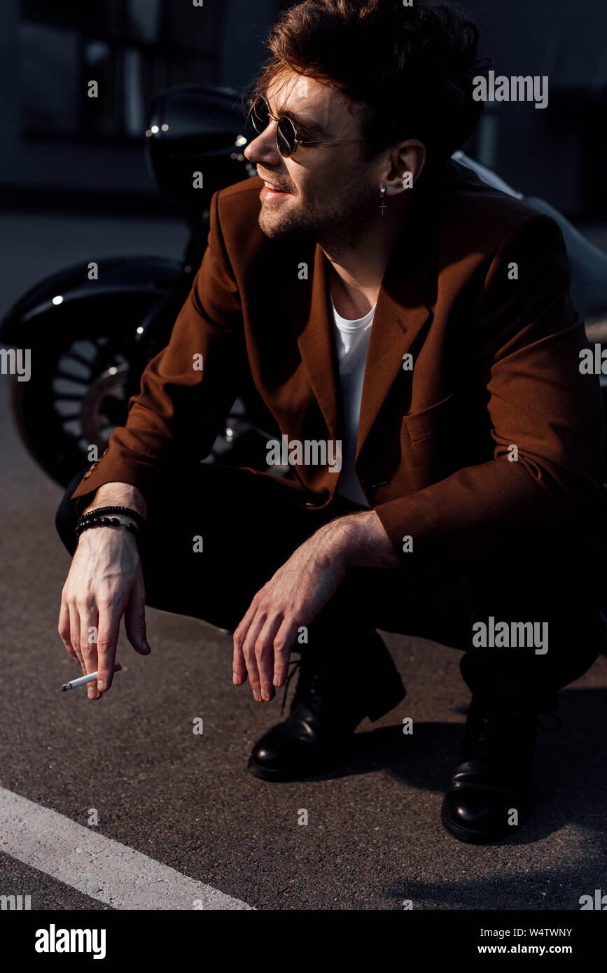 young man in sunglasses and jacket sitting squatting near motorcycle ...