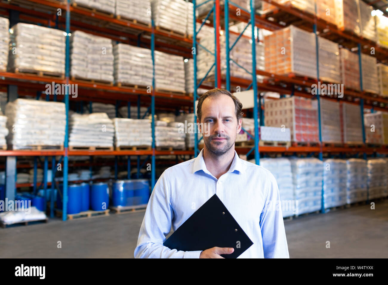Male supervisor holding clipboard and looking at camera in warehouse ...