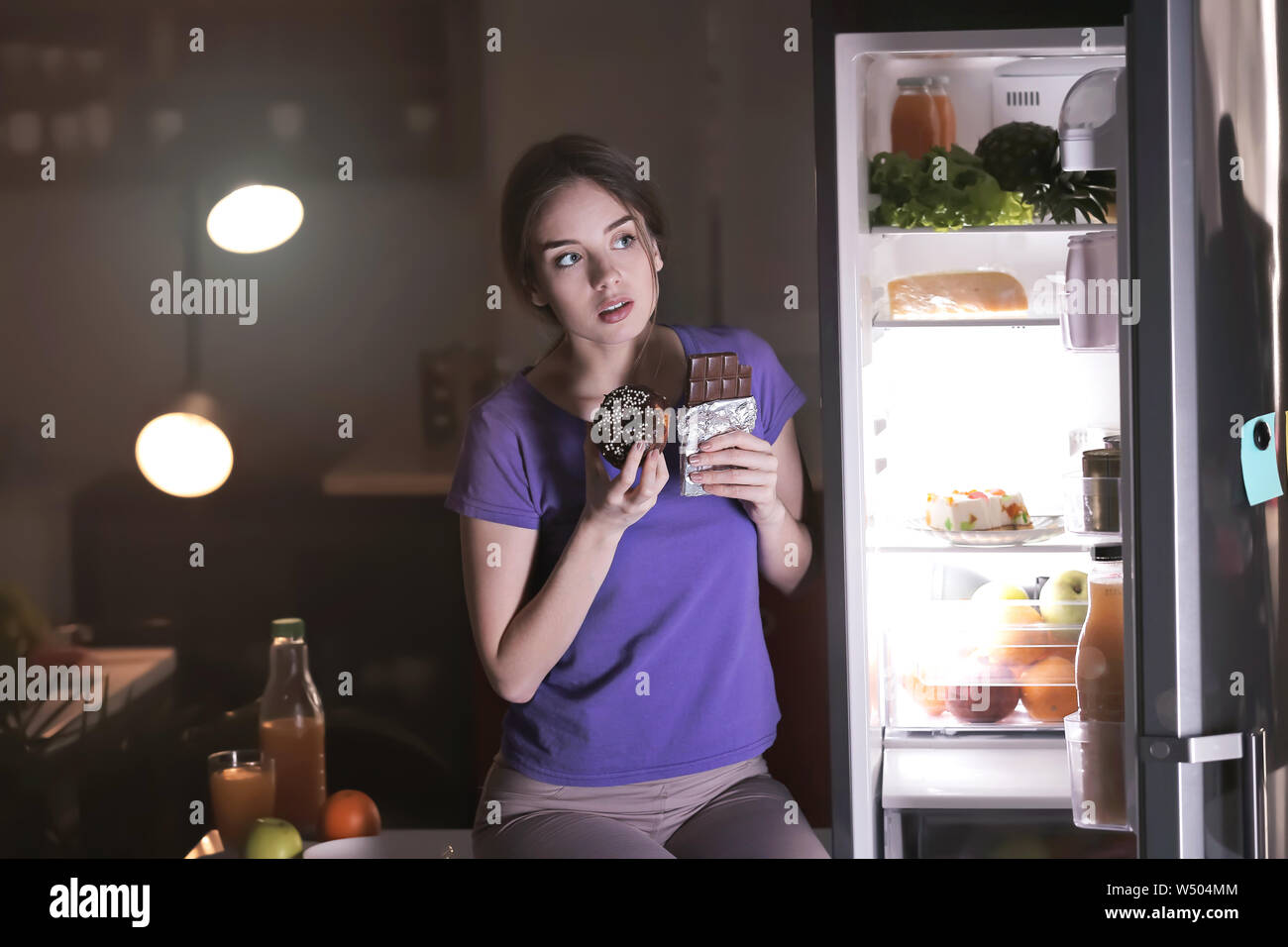 Wary young woman eating unhealthy food near fridge in kitchen at night ...