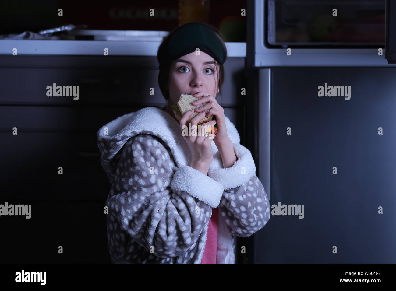 Wary young woman eating unhealthy food near fridge in kitchen at night ...