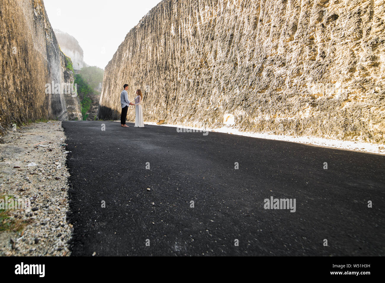 Newlyweds standing among two cliffs on Balinese beach and holding hands ...
