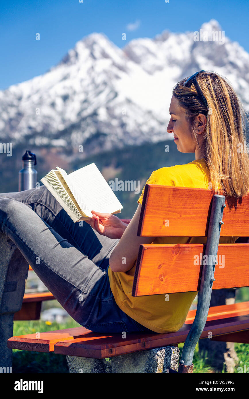 Young beautiful and happy girl reading a book on a bench, mountain ...